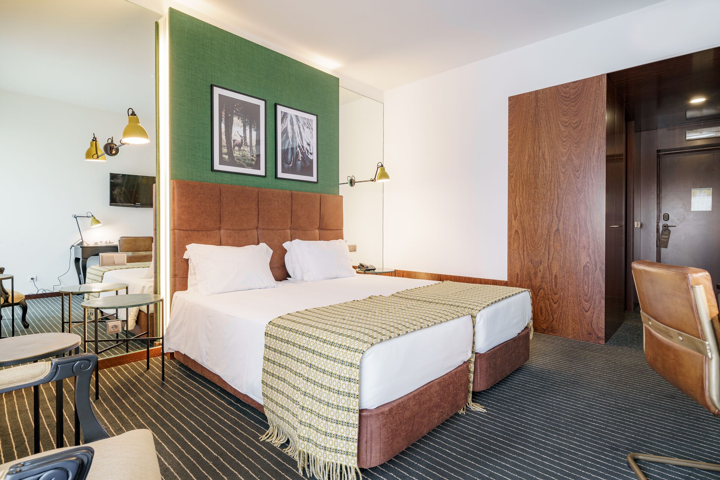 Quarto de hotel com uma cama grande, uma escrivaninha, duas cadeiras, uma parede verde e uma porta de madeira.