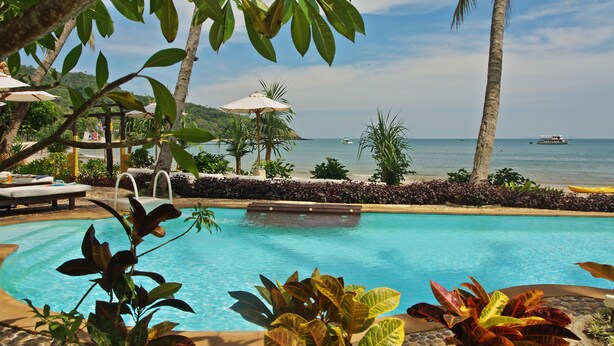 Área de piscina com espreguiçadeiras e guarda-sóis, cercada por plantas tropicais e vista para o mar.