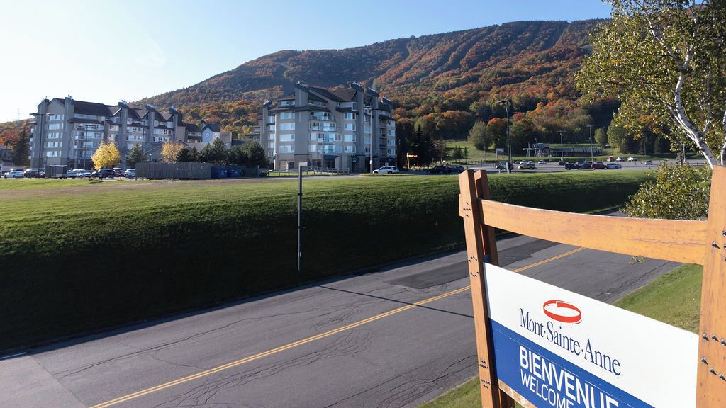 Placa de boas-vindas aos visitantes em Mont Sainte-Anne, com vista para um edifício de vários andares e uma paisagem montanhosa ao fundo.