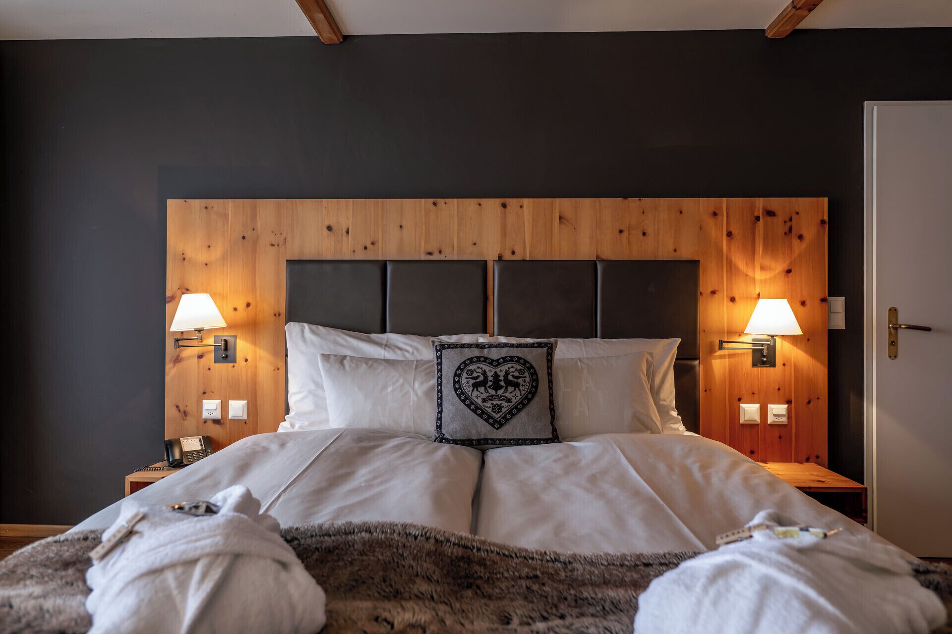 Quarto com cabeceira de madeira, duas luminárias de apoio, um travesseiro em formato de coração e um tapete fofinho.
