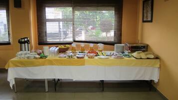 Uma mesa de buffet com diversos alimentos e bebidas, em frente a uma parede amarela com uma janela ao fundo.