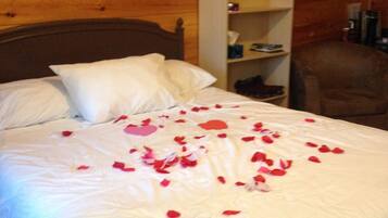 Cama com lençóis brancos e pétalas de rosa espalhadas, cabeceira de madeira, estante branca e uma cadeira em um quarto com paredes de madeira.