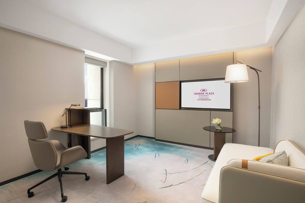 Quarto de hotel moderno com escrivaninha, cadeira de escritório, sofá e tela exibindo 'CROWN PLAZA'.
