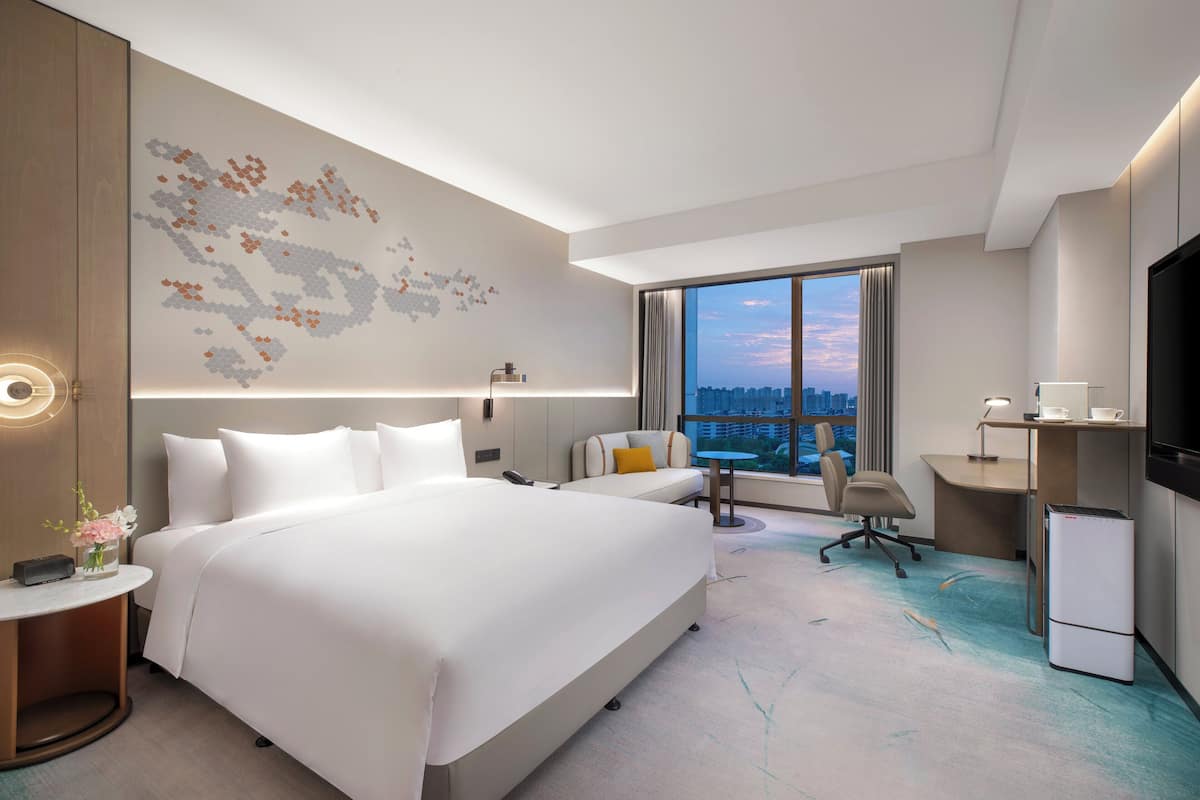 Quarto de hotel moderno com uma cama grande, uma escrivaninha com cadeira, uma mesinha de centro com abajur e uma televisão.