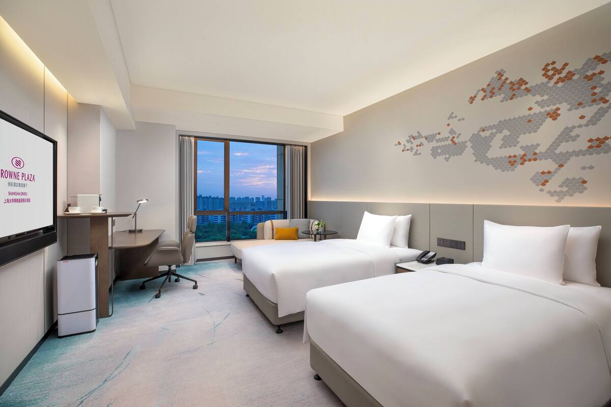 Quarto de hotel com duas camas, uma escrivaninha, uma cadeira, uma mesinha e uma janela grande com vista para a cidade.