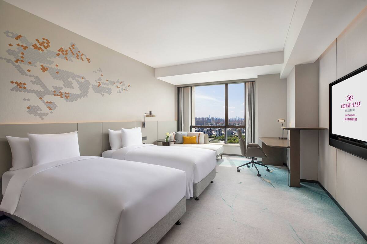 Quarto de hotel com duas camas, uma escrivaninha e uma janela grande com vista para a cidade.