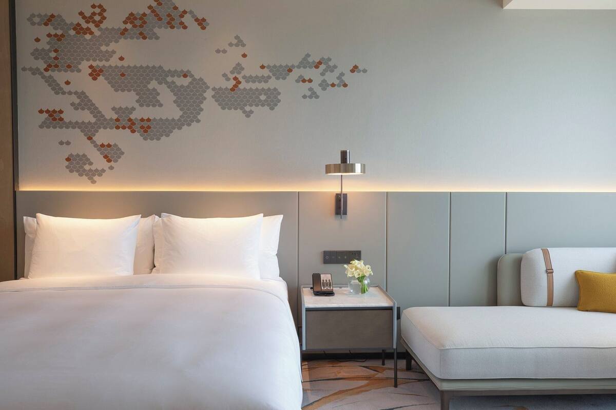 Quarto de hotel com uma cama grande, uma mesa de cabeceira com luminária, uma mesinha com um vaso de flores e um sofá com almofadas.
