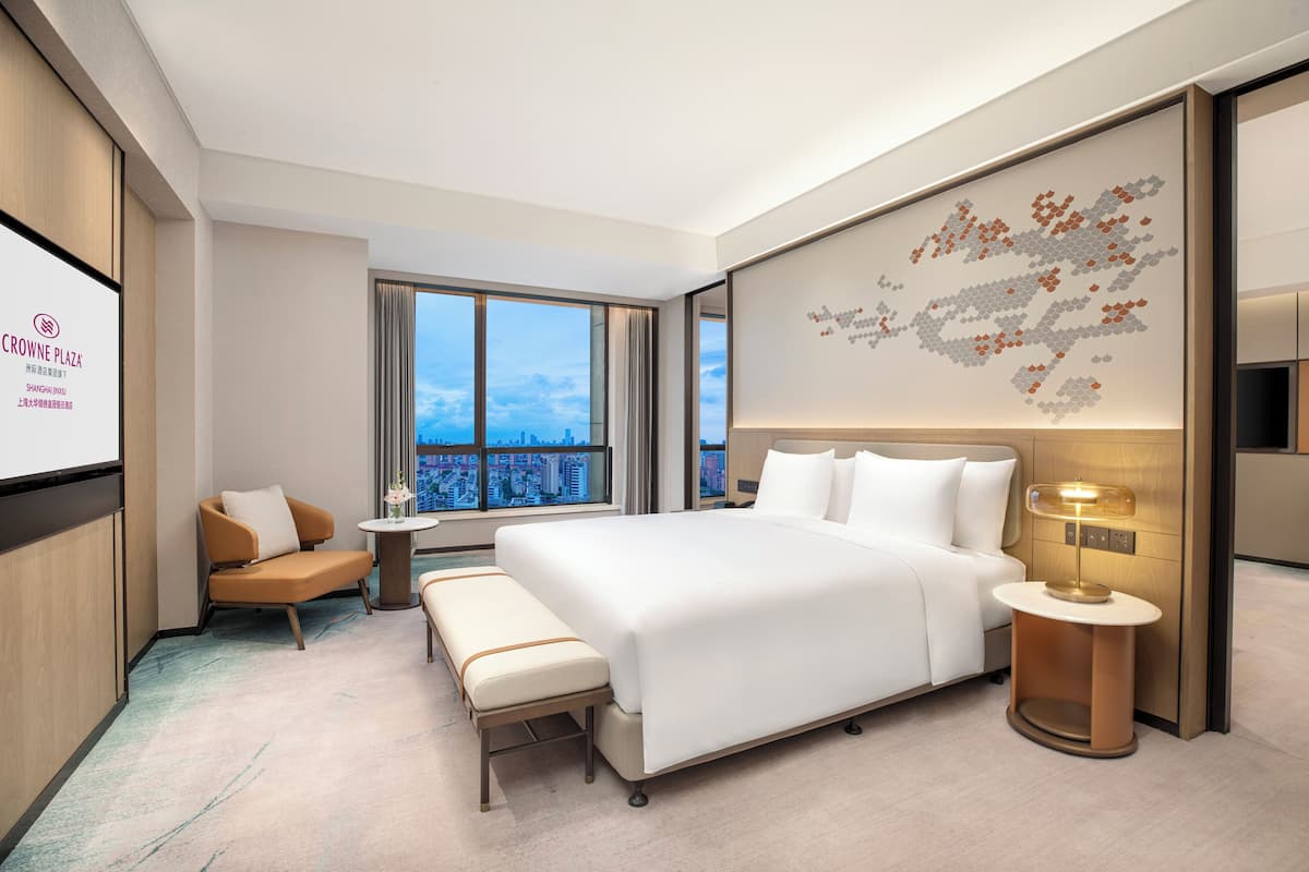 Quarto de hotel moderno com uma cama grande, área de estar com um banco, uma mesa pequena e vista para a cidade através de amplas janelas.