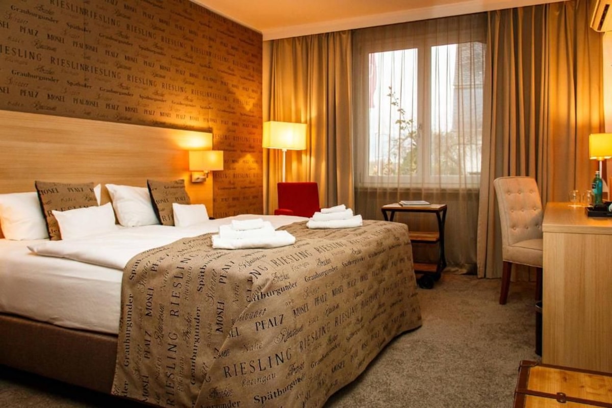 Quarto de hotel com uma cama, escrivaninha, cadeira e janela com cortinas.