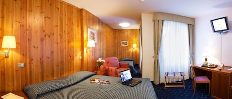 Quarto de hotel com parede revestida em madeira, cama com roupa de cama estampada, escrivaninha com cadeira, televisão fixada na parede e janela com cortinas.