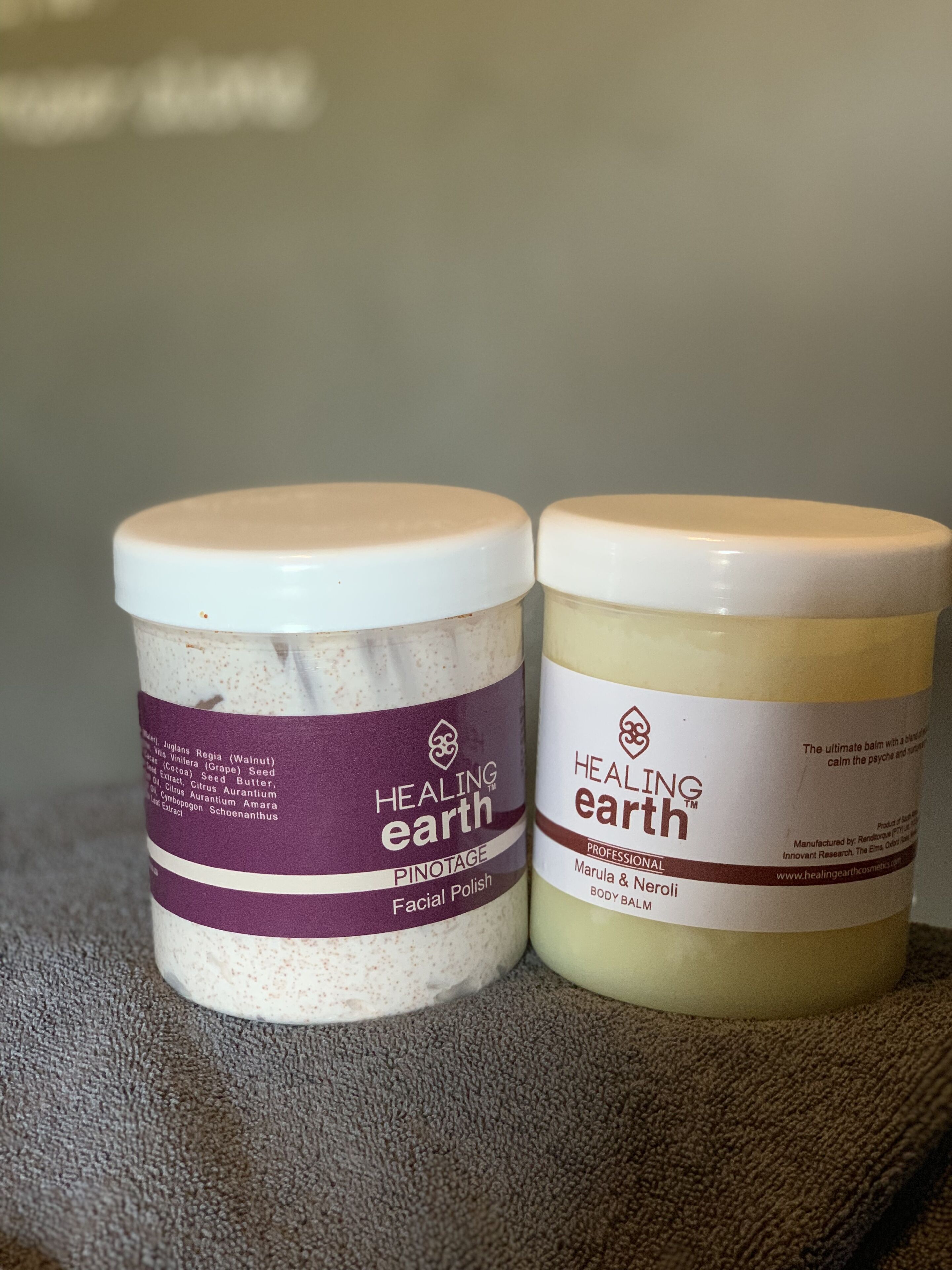 Dois produtos da Healing Earth, um identificado como esfoliante facial e outro como bálsamo corporal, sobre uma superfície texturizada.