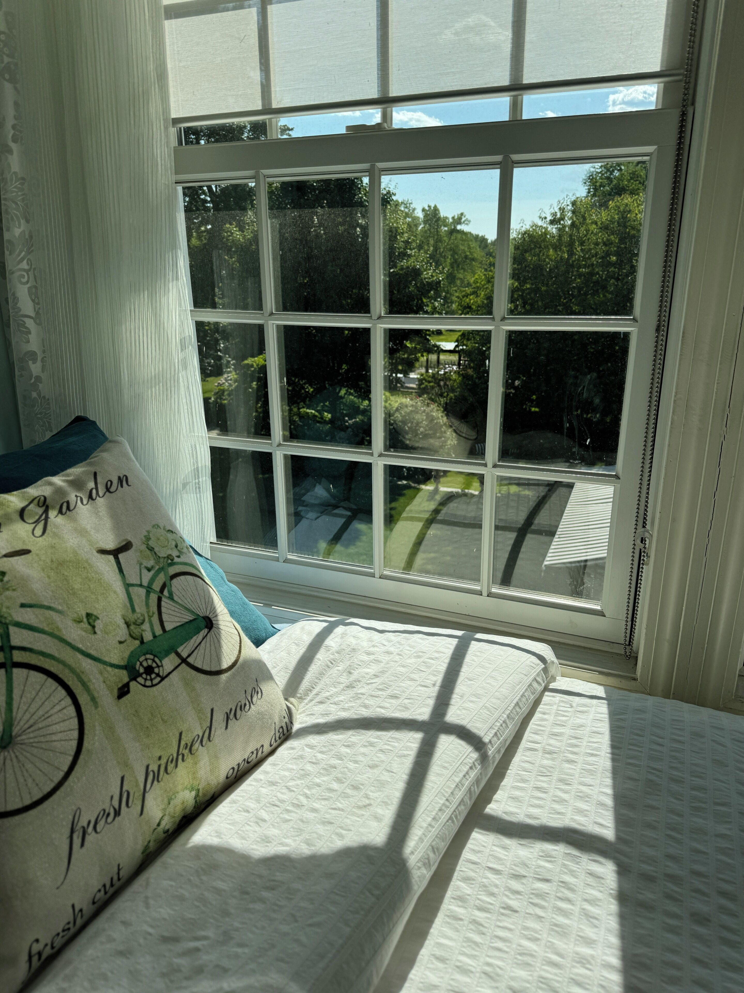 Uma janela com vista para um jardim, um travesseiro com desenho de bicicleta e uma cama com cobertura branca.