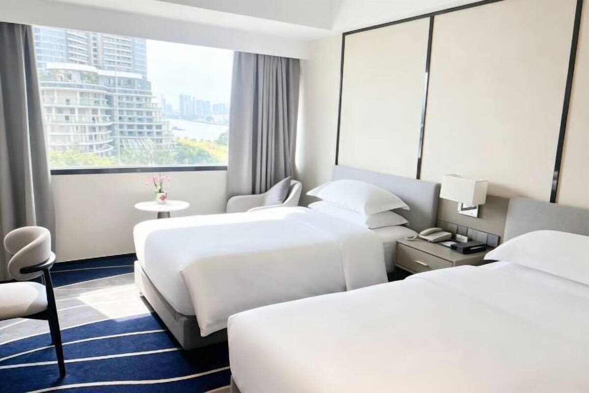 Quarto de hotel com duas camas, uma mesinha, uma cadeira, mesa de cabeceira com abajur e vista para a cidade.