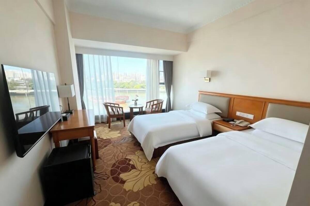 Quarto de hotel com duas camas, uma escrivaninha, uma cadeira, uma televisão e uma janela com vista para a cidade.