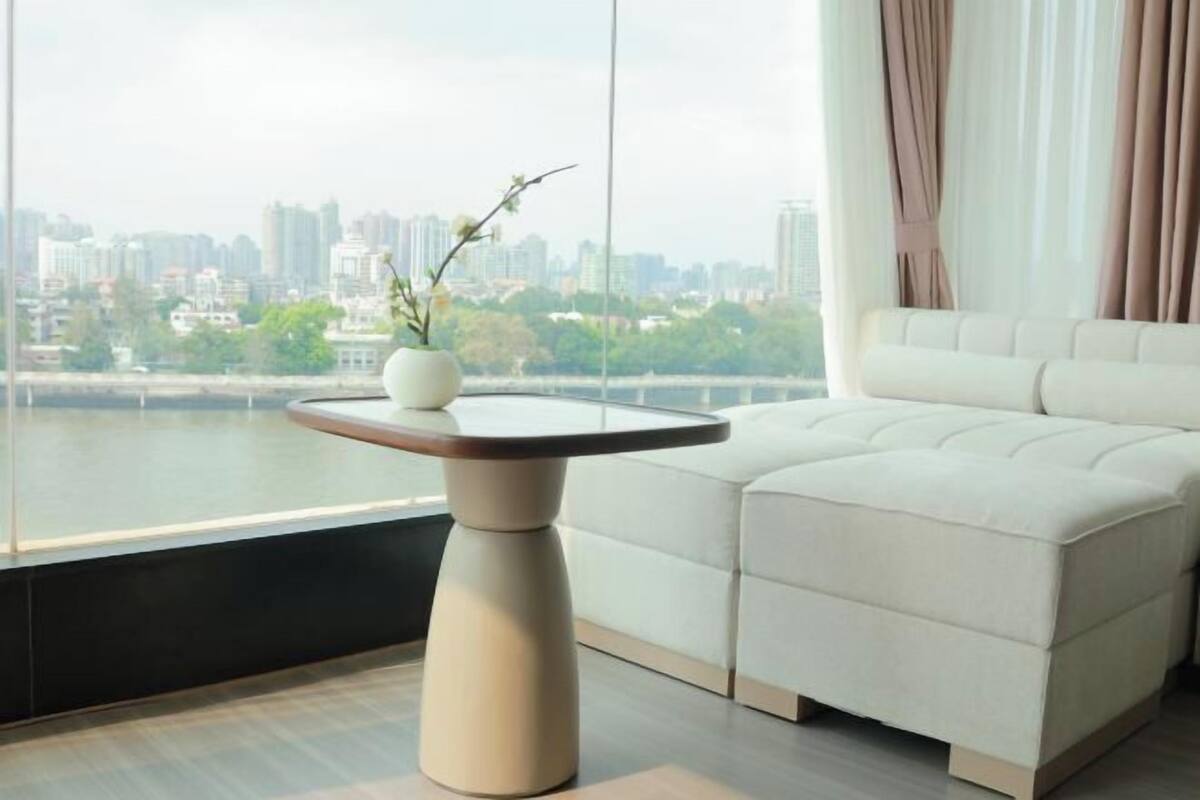 Uma sala de estar moderna com um sofá branco, uma mesa redonda com um vaso de flores e uma janela grande com vista para a cidade.