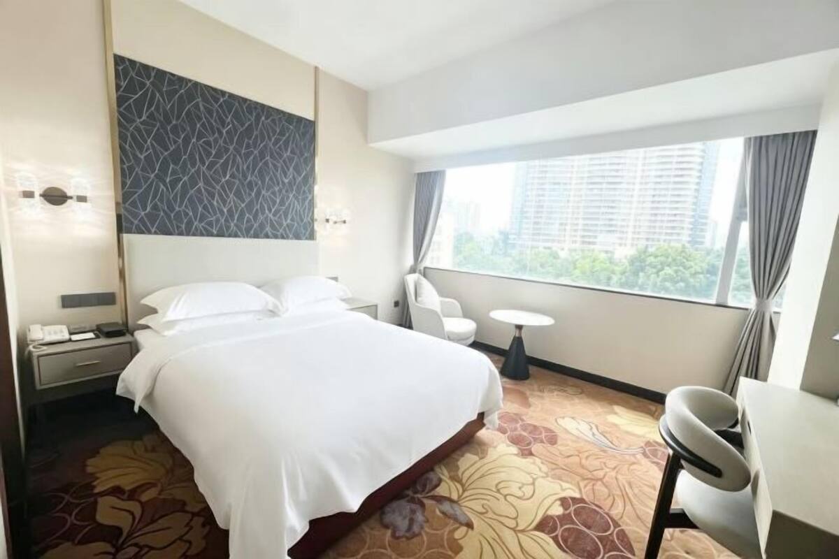 Quarto de hotel com cama, escrivaninha, cadeira, mesa pequena e janela com cortinas.