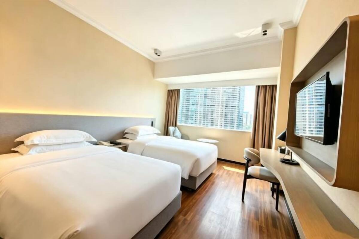 Quarto de hotel com duas camas, uma mesa, uma cadeira, uma televisão e uma janela com cortinas.