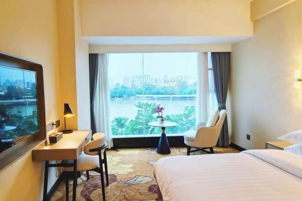 Quarto de hotel com uma cama, uma mesa com cadeira, uma mesinha com um vaso, e uma janela grande com vista para um rio e edifícios.