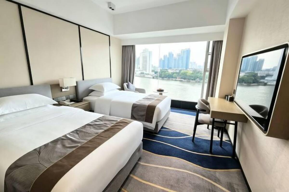Quarto de hotel com duas camas, uma escrivaninha e uma janela grande com vista para a cidade.
