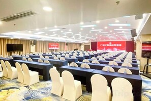 Uma sala de conferências com mesas e cadeiras dispostas em fileiras, uma tela grande e um banner vermelho.