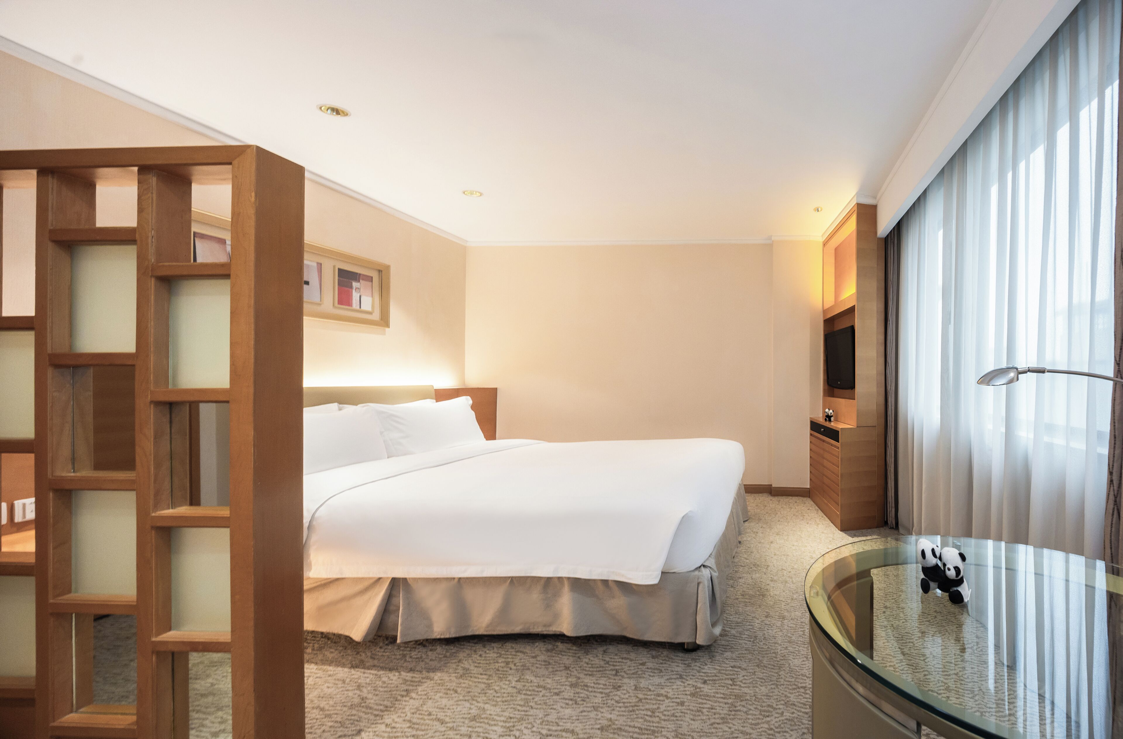 Quarto de hotel com cama, mesa de vidro com um brinquedo, prateleira de madeira e televisão na parede.