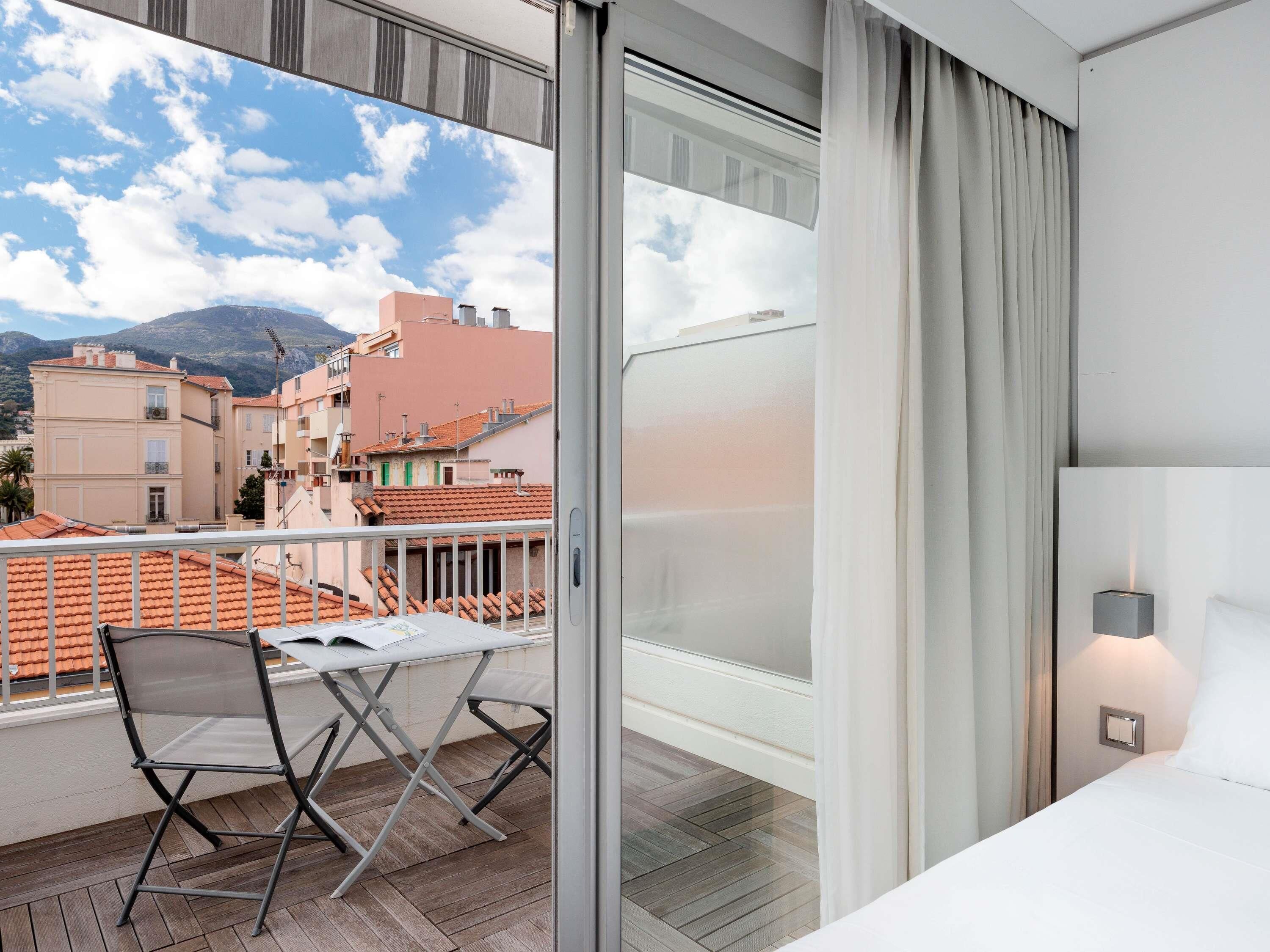 Varanda com mesa e cadeiras, cama com mesa de cabeceira e vista para edifícios e montanhas.