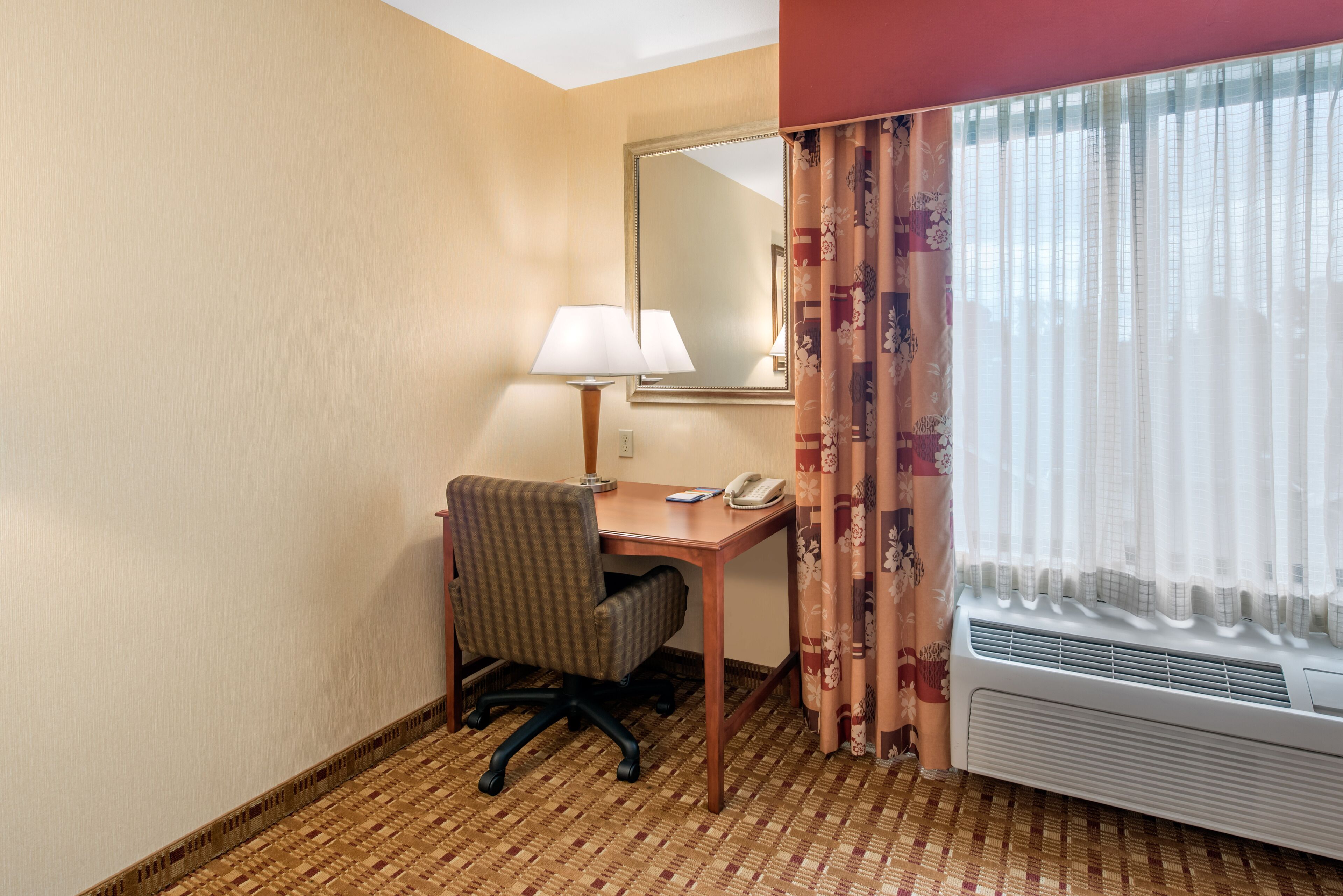 Quarto de hotel com escrivaninha, cadeira, luminária, telefone e ar-condicionado.