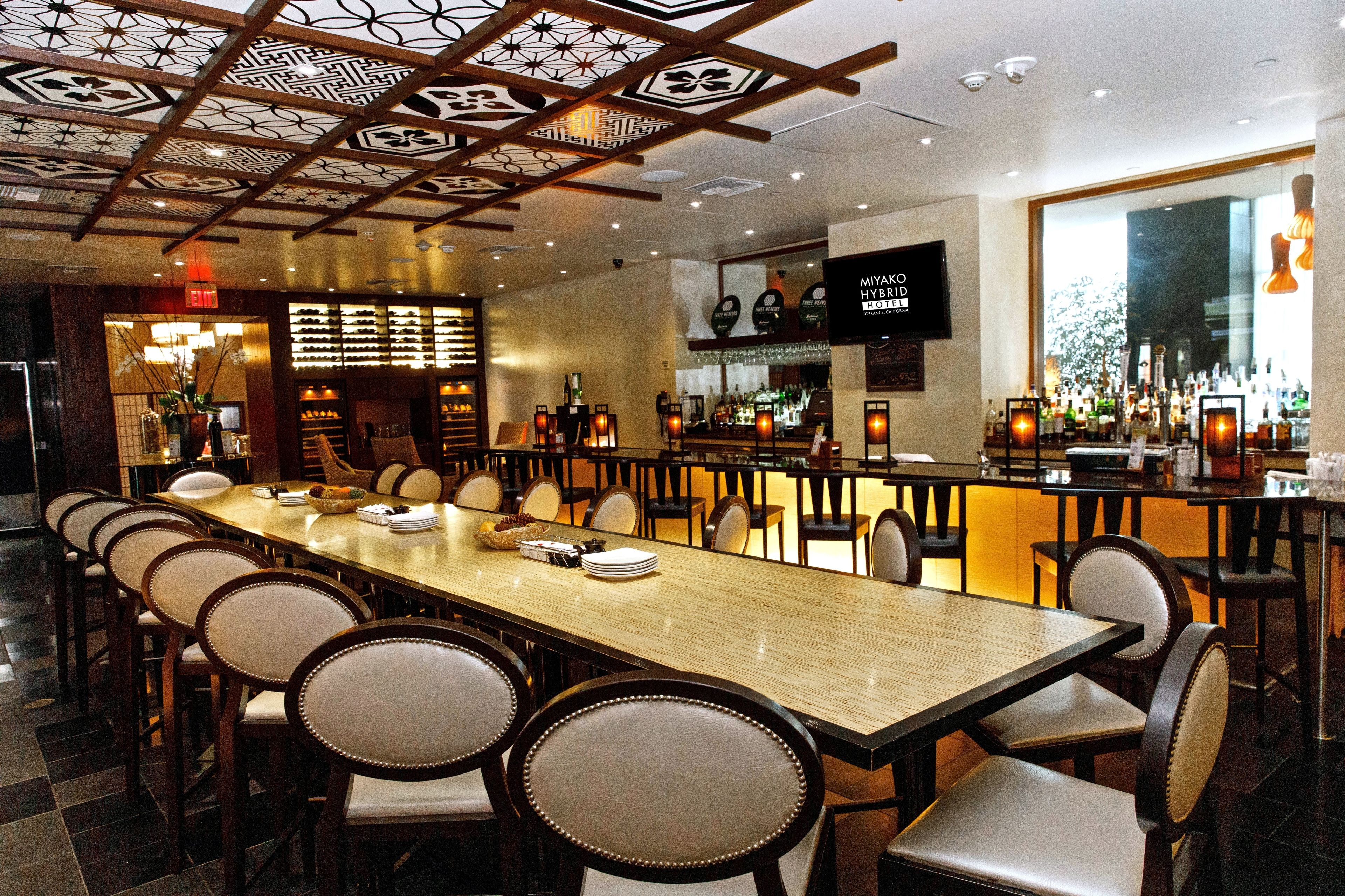 Um restaurante com uma longa mesa de madeira e várias cadeiras, um bar com garrafas, e uma televisão na parede.