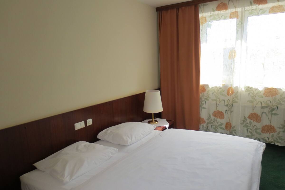 Cama bem arrumada com lençóis brancos, luminária de cabeceira e cortina florida.