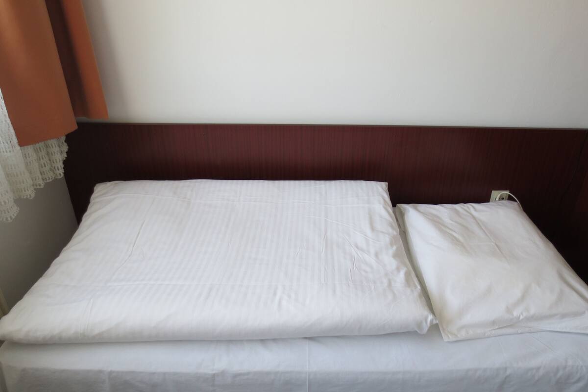 Cama com lençóis brancos e cabeceira de madeira em quarto com paredes simples.