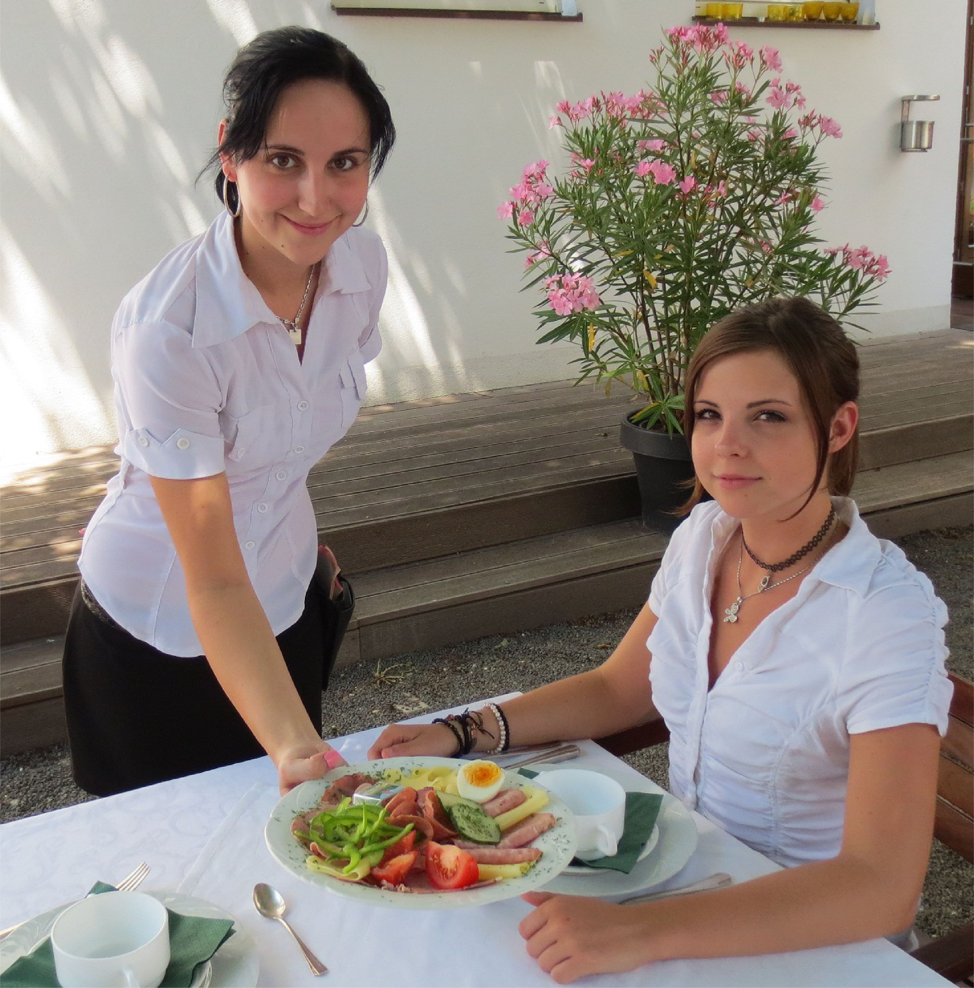 Duas mulheres sentadas à mesa num espaço ao ar livre, com uma refeição, rodeadas por uma planta em vaso com flores cor-de-rosa.