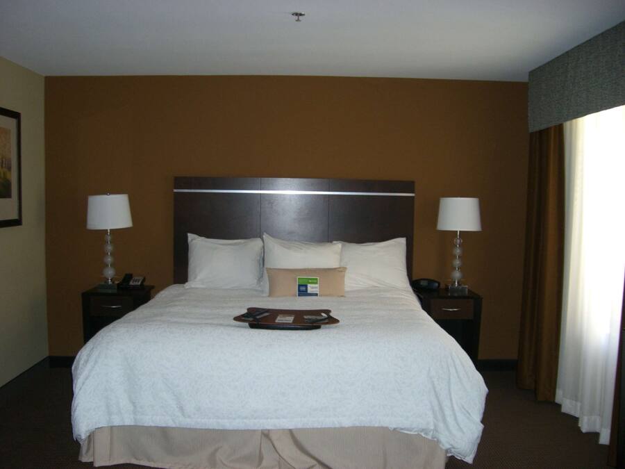 Quarto de hotel com uma cama grande, duas mesas de cabeceira com luminárias, uma mesinha de apoio e uma janela com cortinas.