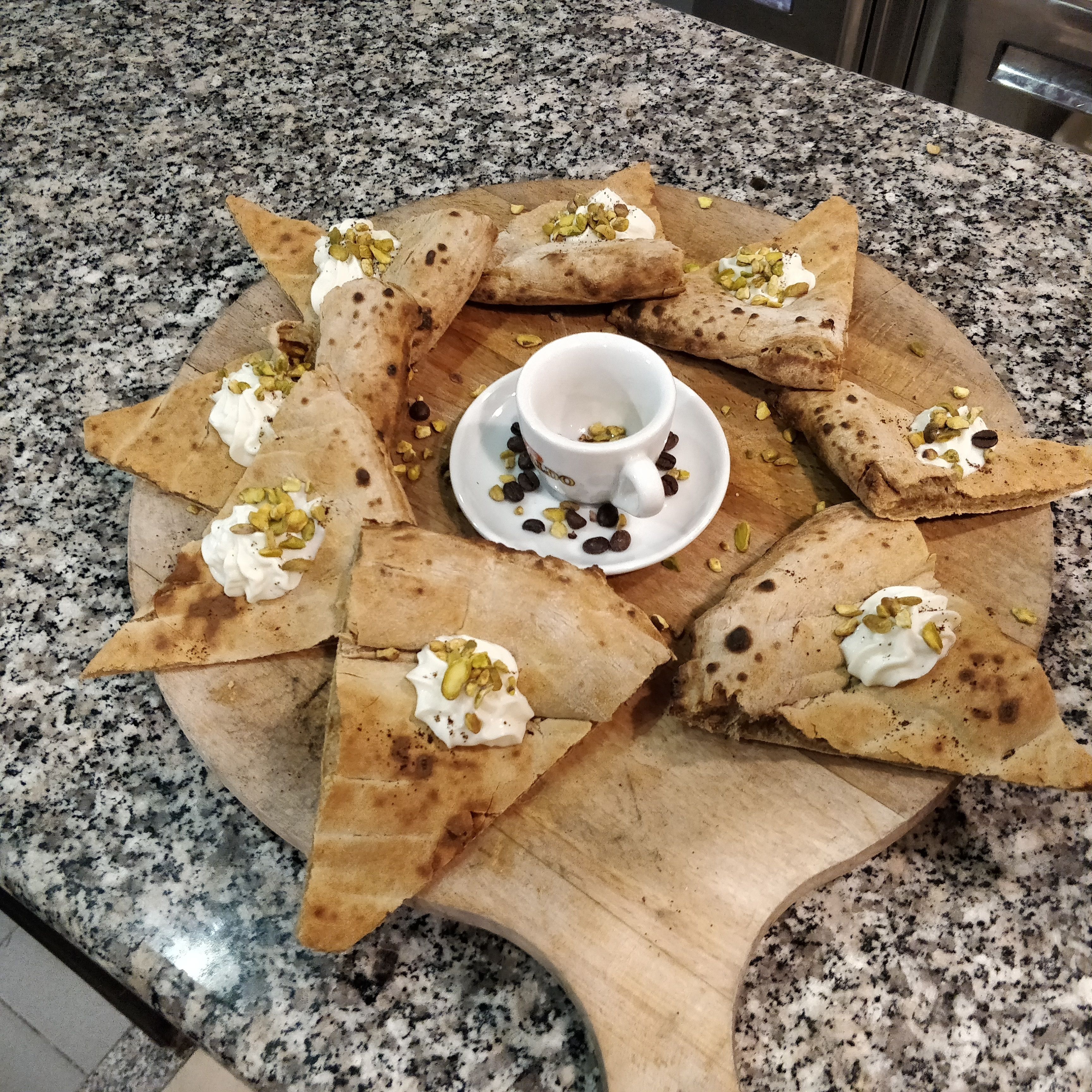 Uma tábua de madeira com doces triangulares cobertos de chantilly e pistache, acompanhada de uma pequena xícara de café.