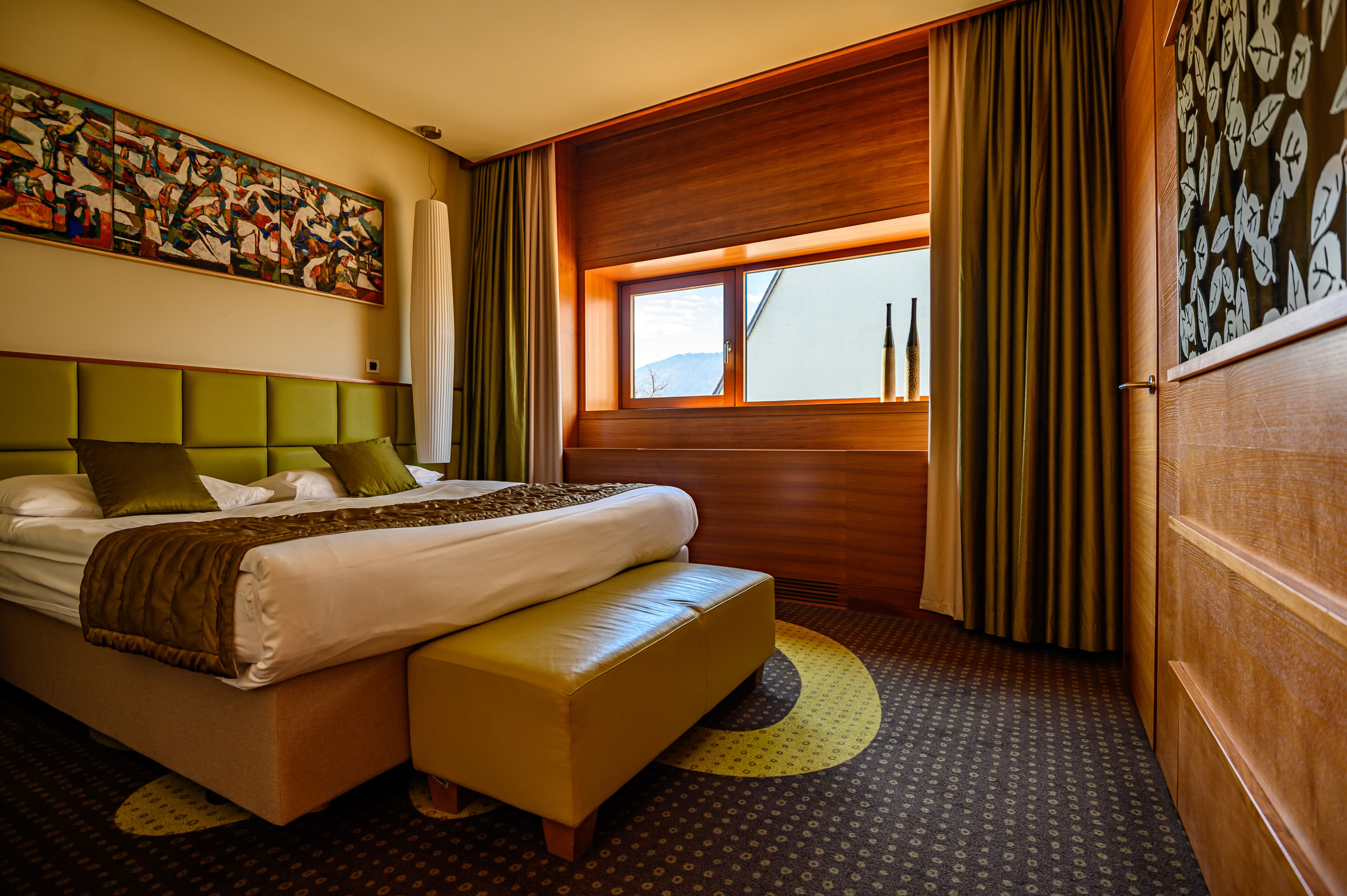 Quarto de hotel com uma cama grande, um banco, janela com cortinas, uma obra de arte na parede e tapete estampado.