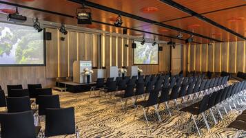 Uma sala de conferências com fileiras de cadeiras, uma tela grande e um púlpito.