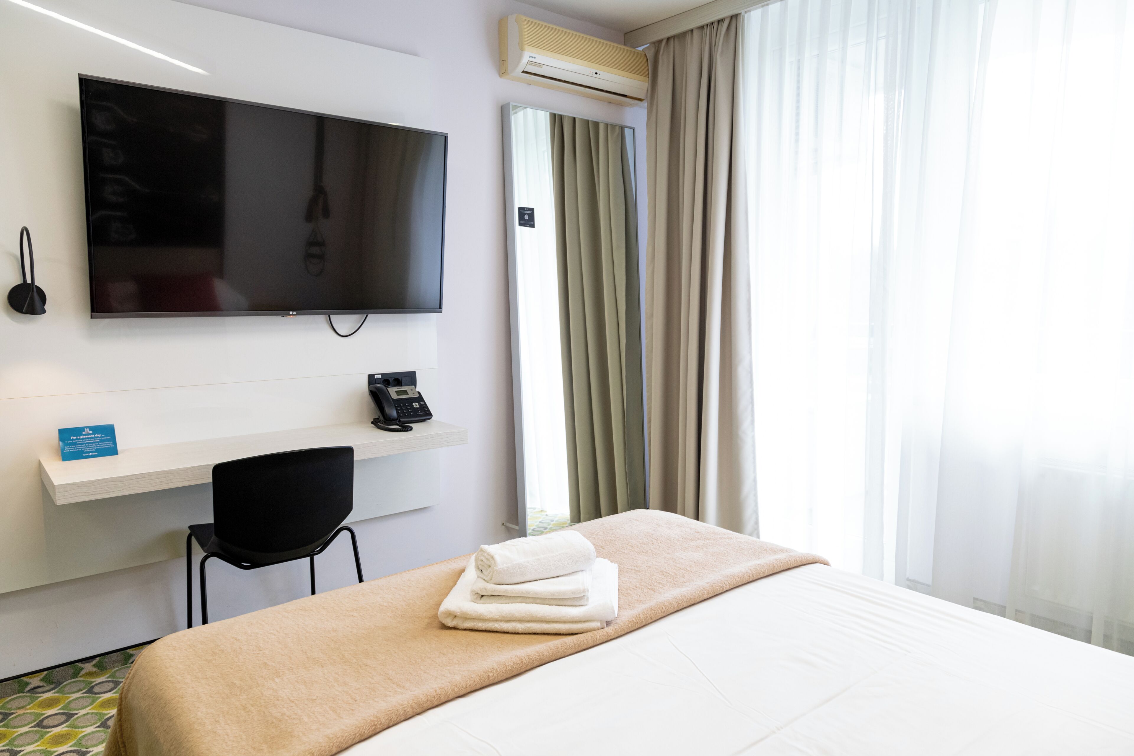 Quarto de hotel com cama, televisão, telefone e janela com cortinas.