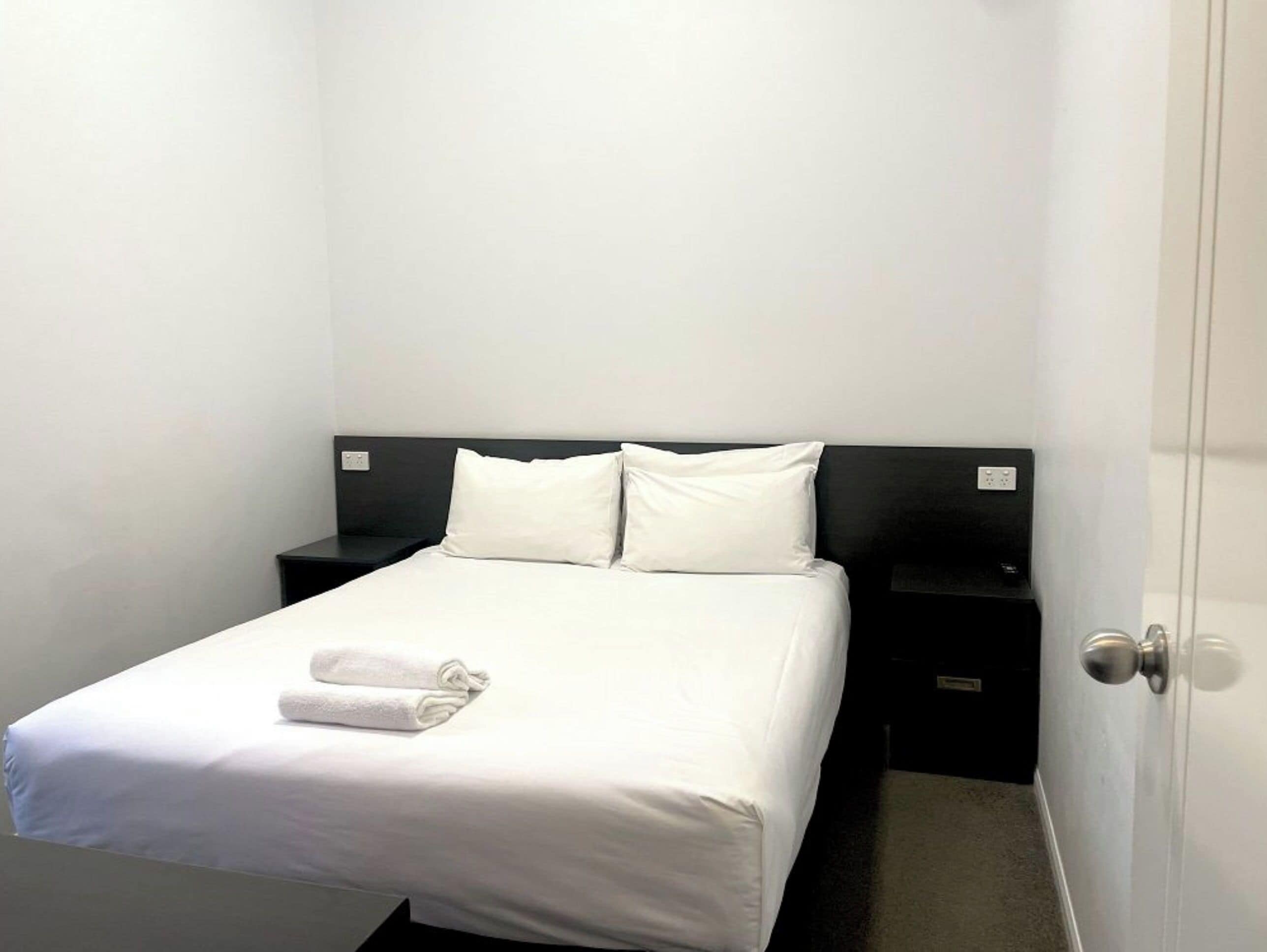 Cama bem arrumada com roupa de cama e almofadas brancas, toalha dobrada sobre a cama e mesa de cabeceira preta com tomada.