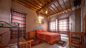 Um quarto com uma cama, um banco de madeira, uma mesa de cabeceira, um abajur e uma janela com cortinas listradas.