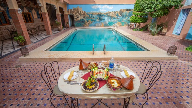 Área de refeições à beira da piscina, com uma mesa posta para dois, cadeiras ao redor e vista para a piscina.
