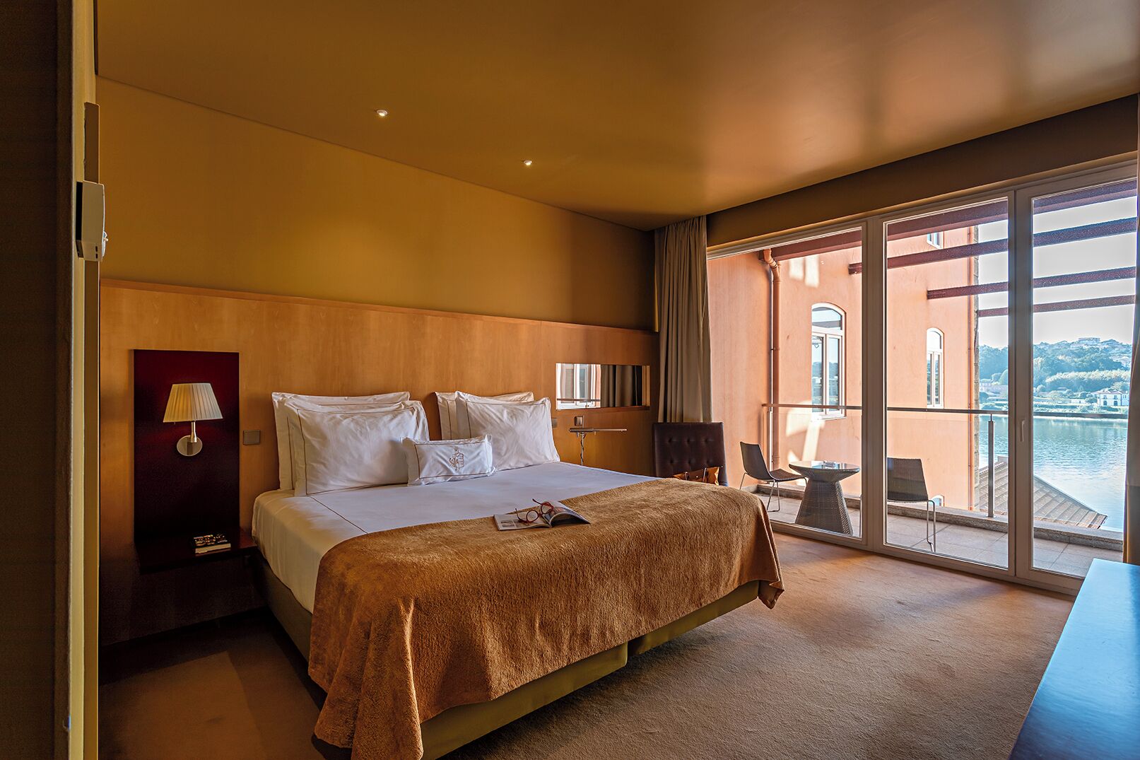 Quarto de hotel com uma cama grande, mesa de cabeceira, luminária, cadeira e varanda com vista para a água.