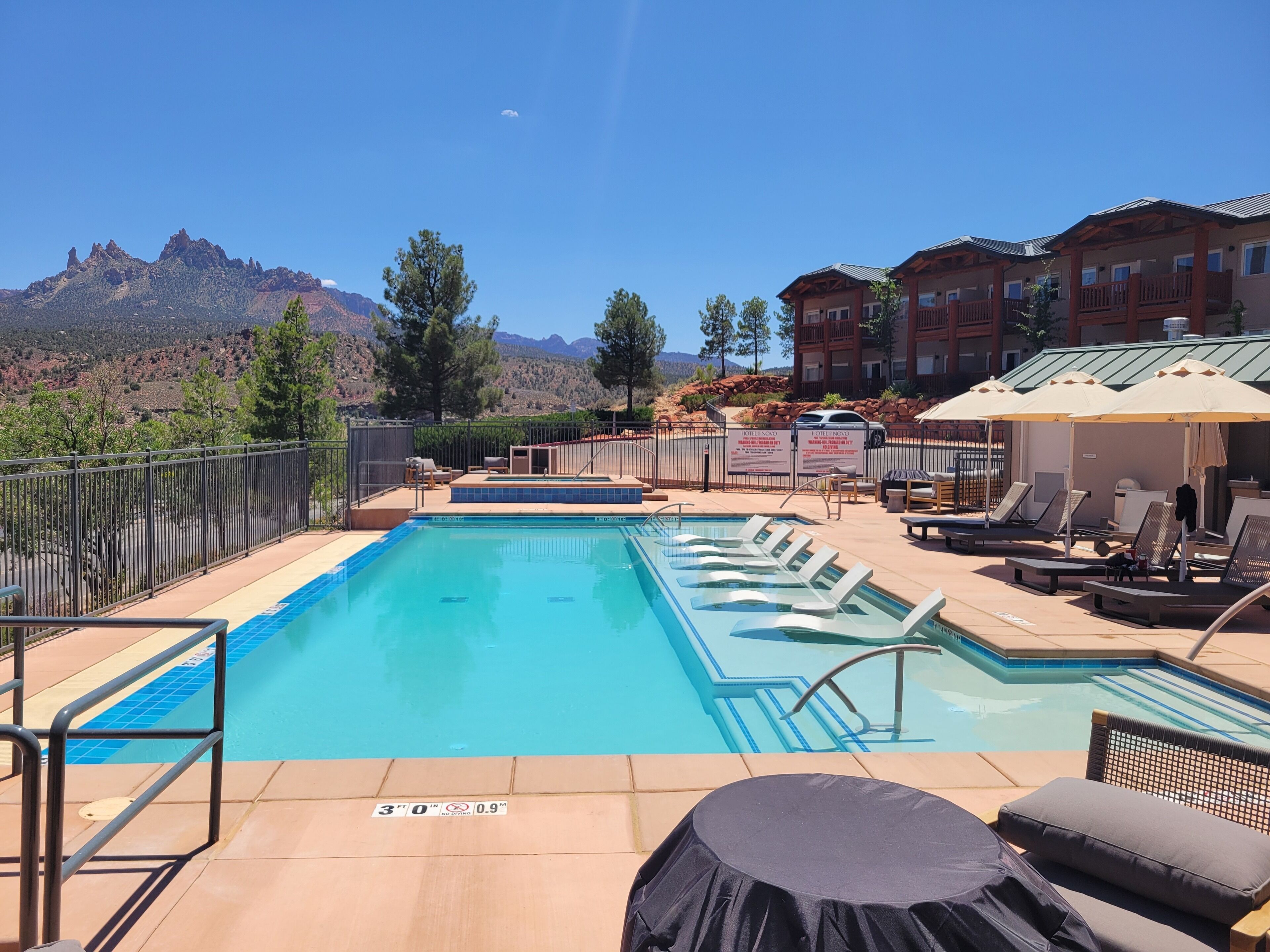 Área de piscina com espreguiçadeiras e guarda-sóis, cercada por uma grade, com montanhas ao fundo.