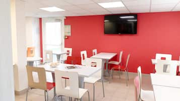 Uma sala de jantar com mesas e cadeiras brancas, parede vermelha e televisão de tela plana na parede.