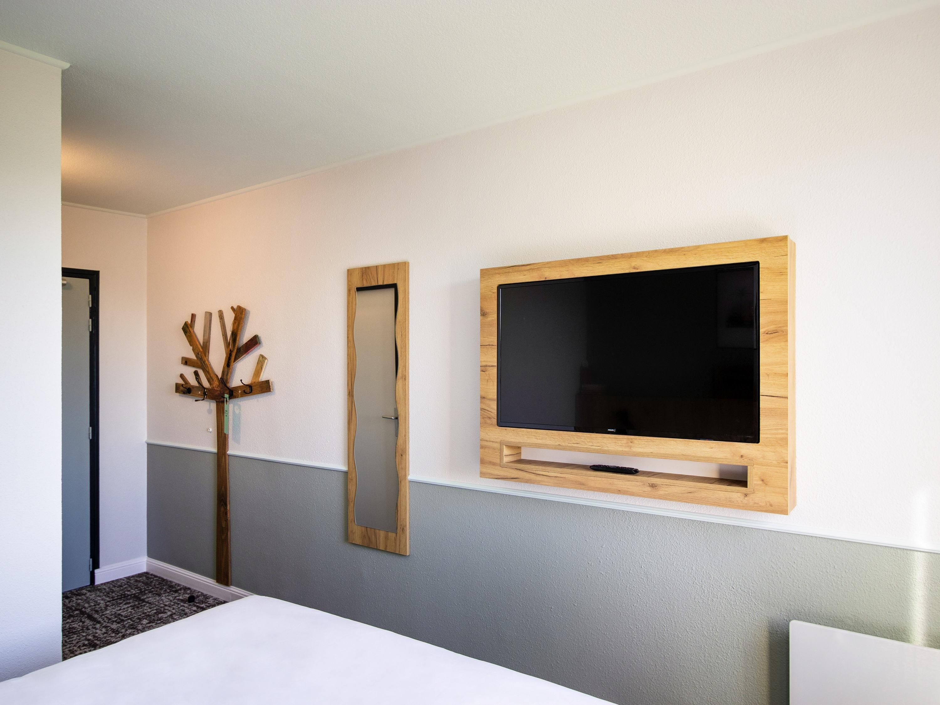 Quarto de hotel com uma cama, um porta-roupas de madeira, um espelho e uma televisão fixada na parede.