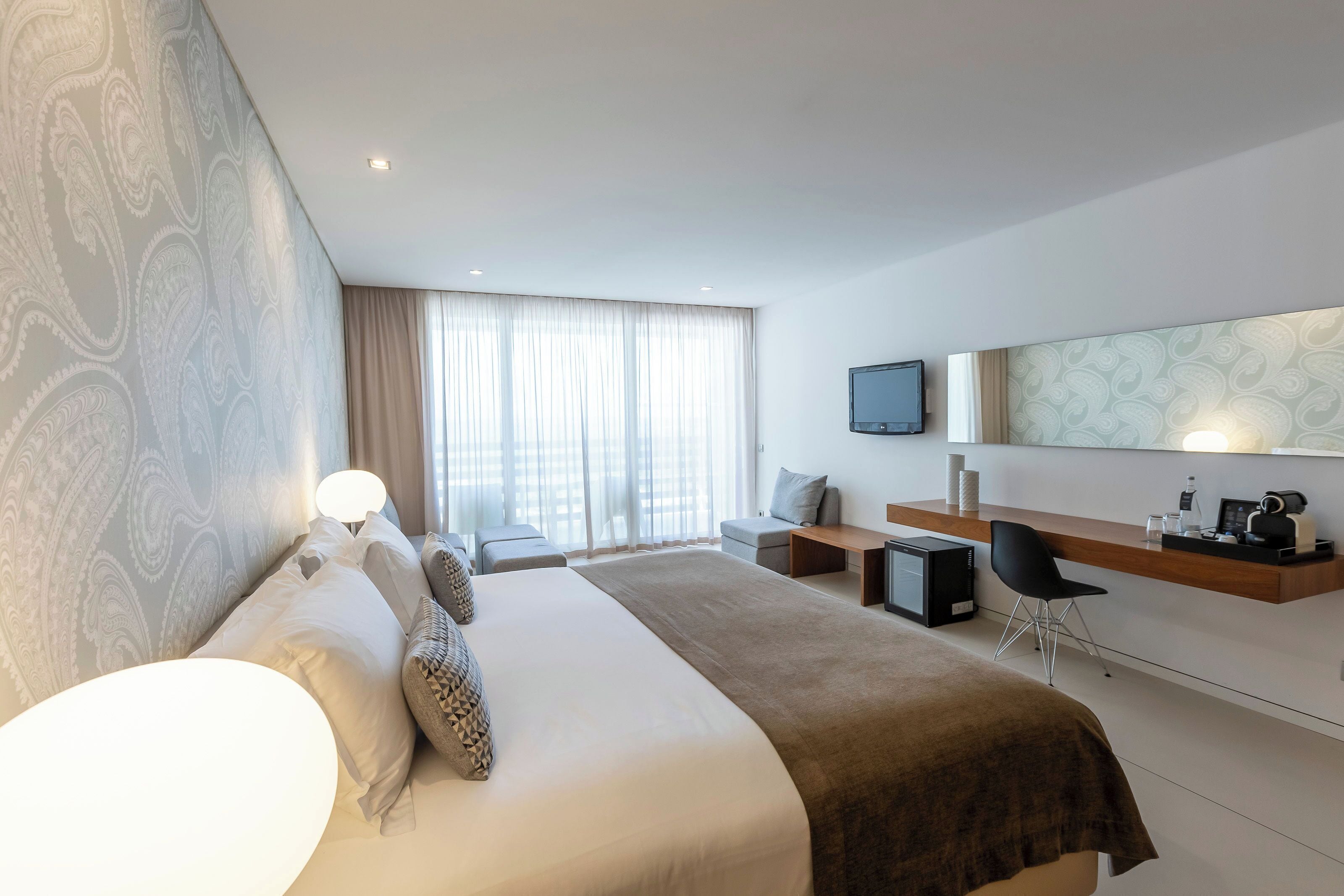 Quarto de hotel moderno com uma cama grande, uma escrivaninha, uma cadeira, uma televisão e um abajur.