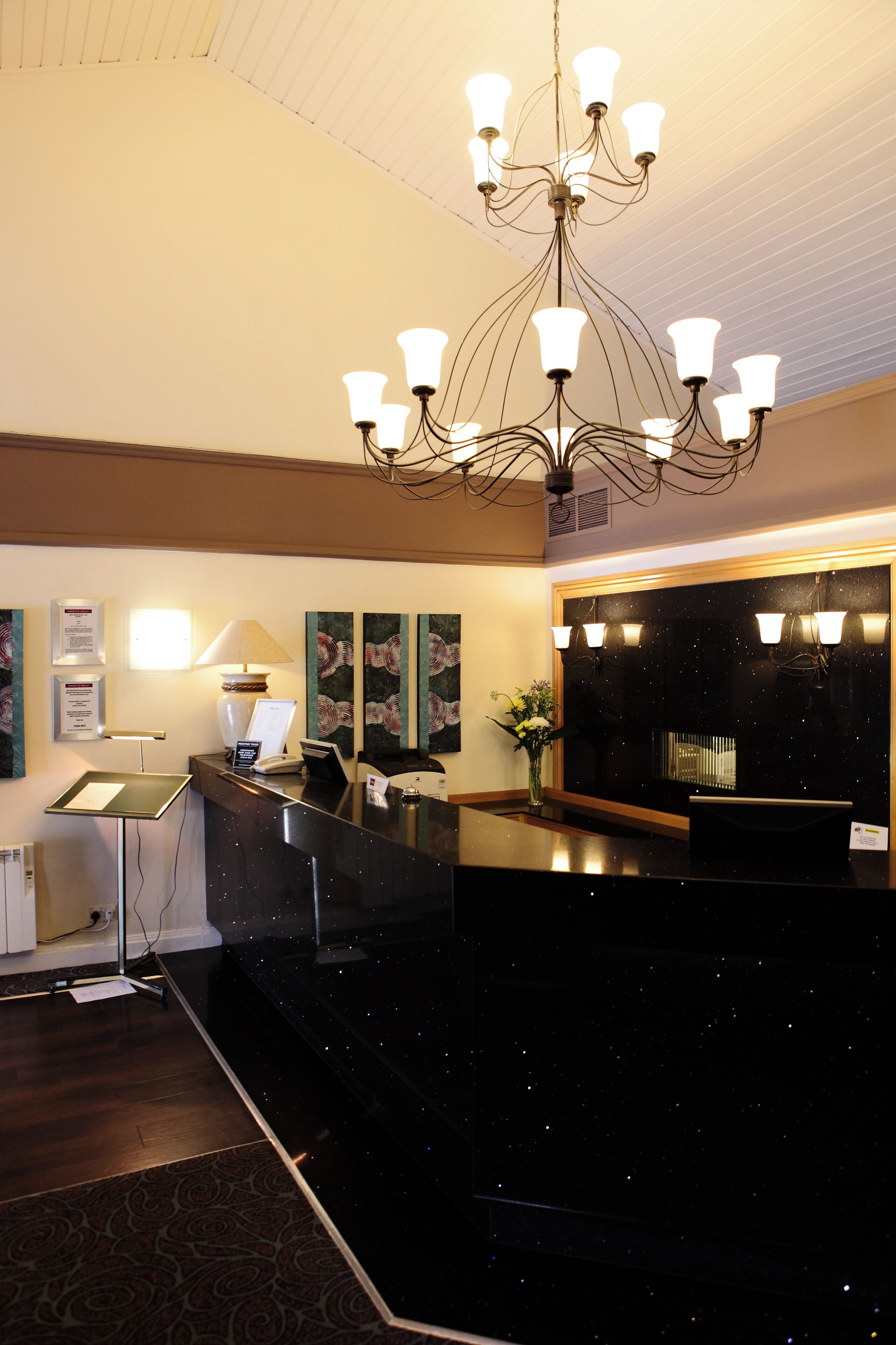 Uma área de recepção de hotel com balcão de mármore preto, um candelabro com várias luminárias, obras de arte emolduradas na parede e um balcão de atendimento com computador e telefone.