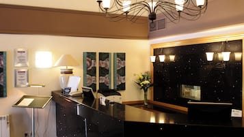 Uma área de recepção de hotel com balcão de mármore preto, um candelabro com várias luminárias, obras de arte emolduradas na parede e um balcão de atendimento com computador e telefone.