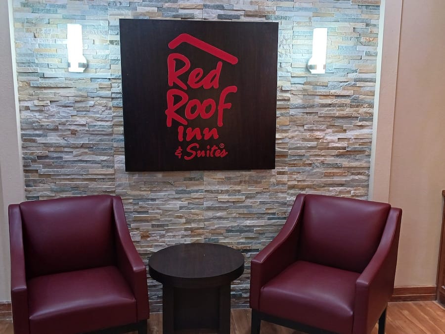 Placa da Red Roof Inn com um muro de pedra ao fundo, duas poltronas de couro vermelho e uma pequena mesa redonda em uma área de recepção.