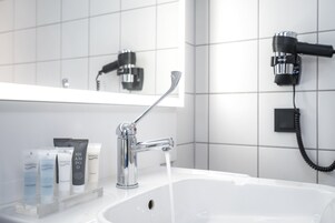 Casa de banho com lavatório branco, torneira cromada e secador de cabelo fixado na parede.