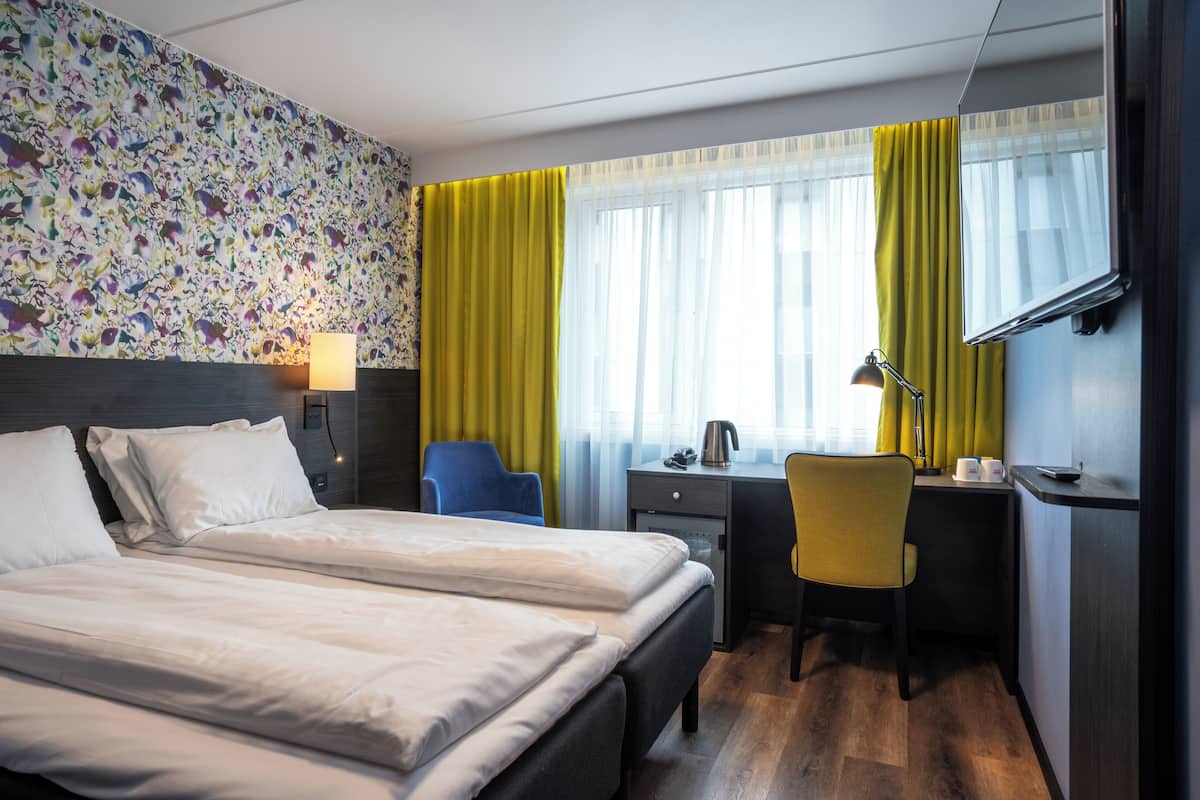 Quarto de hotel com duas camas, uma escrivaninha, uma cadeira, abajur e janela com cortinas amarelas.