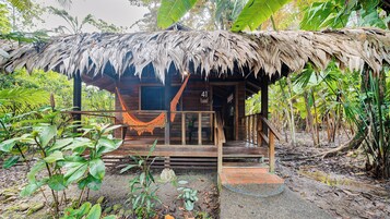 Uma cabana de madeira com telhado de palha, cercada por vegetação exuberante e uma pequena varanda com uma rede.